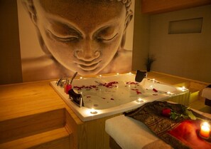 Uma área de spa com uma banheira grande cheia de pétalas de rosa e velas, mobiliário de madeira e uma estátua de Buda na parede.