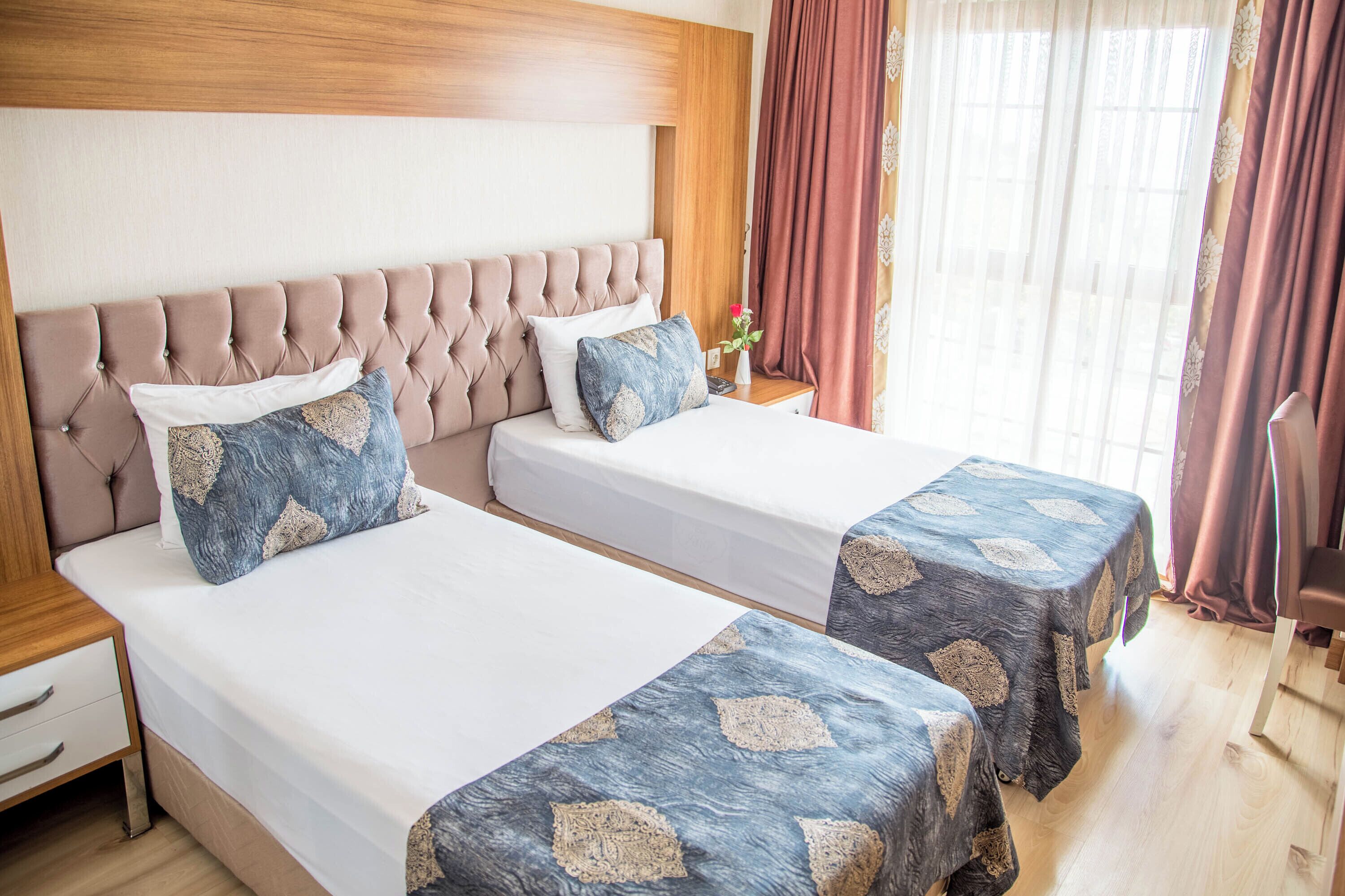 Quarto de hotel com duas camas, uma cabeceira de madeira, uma mesinha de cabeceira e uma janela com cortinas.