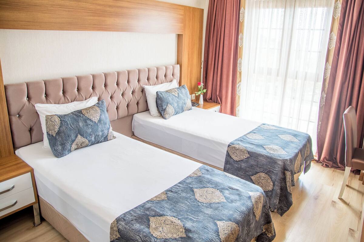 Quarto de hotel com duas camas, uma cabeceira de madeira, uma mesinha de cabeceira e uma janela com cortinas.