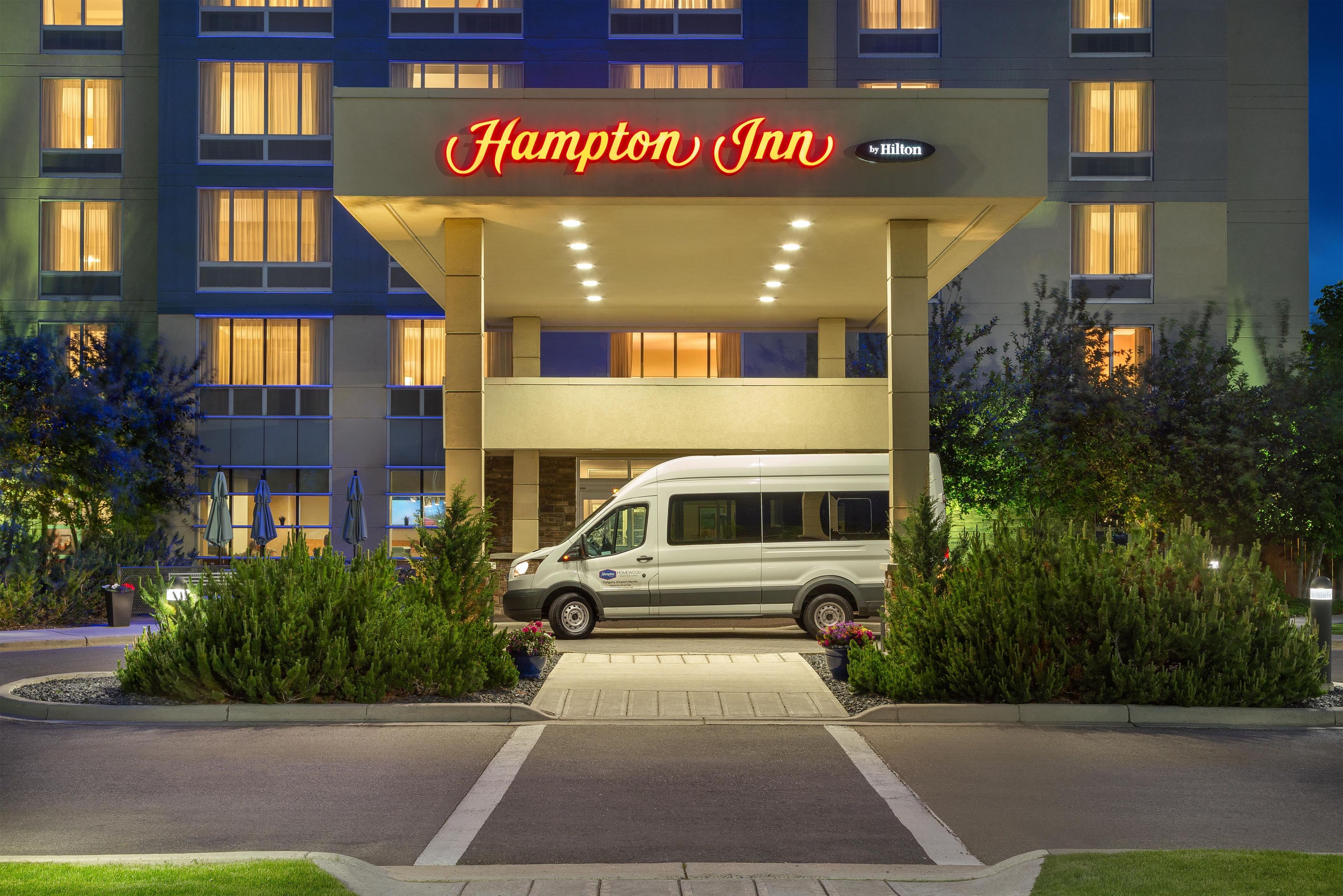 Entrada de um hotel Hampton Inn, com uma van branca estacionada em frente, cercada por vegetação.