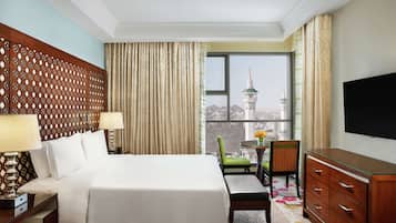Quarto de hotel com uma cama grande, uma mesa com cadeira, televisão e vista para uma mesquita com minarete.