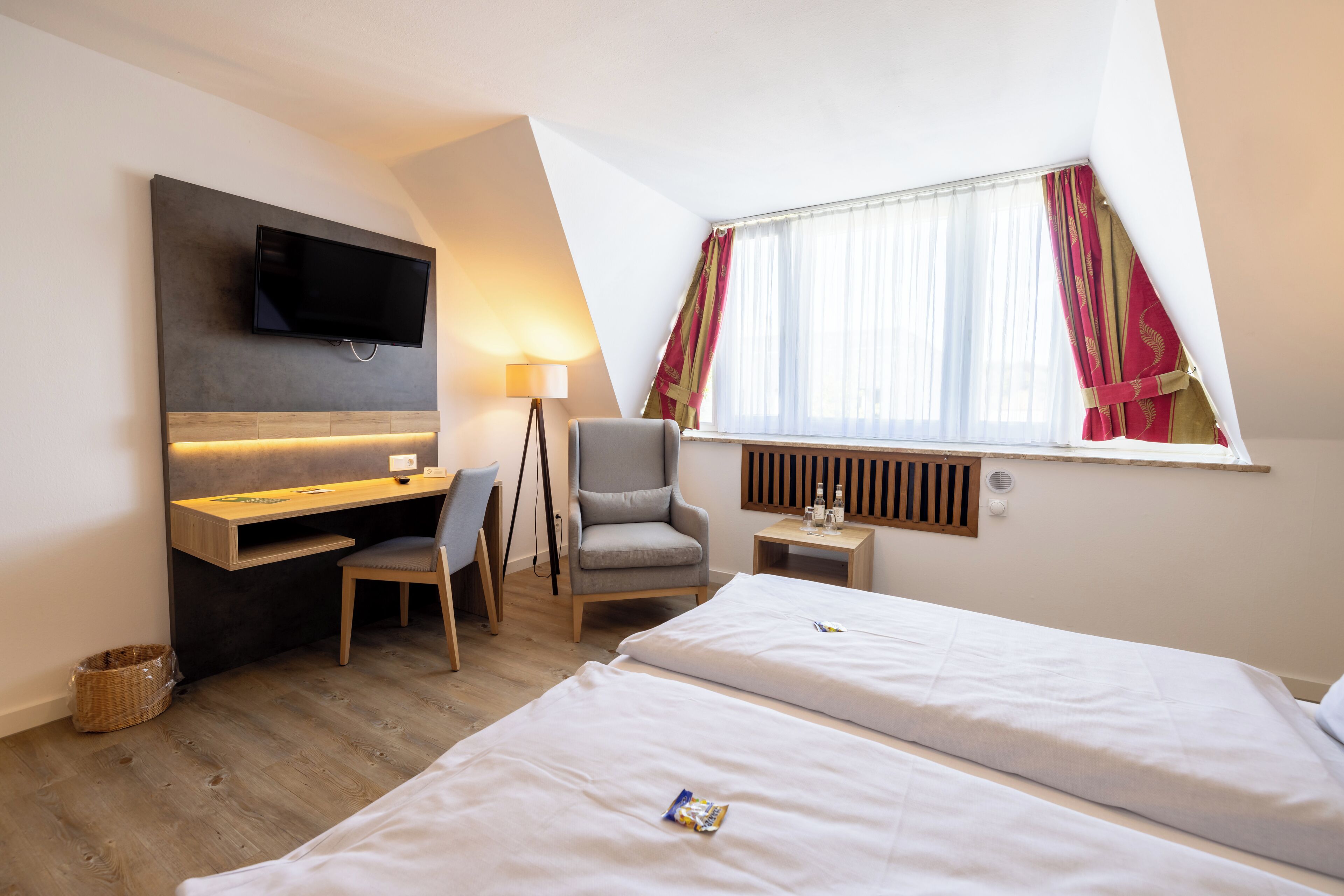 Quarto de hotel com televisão na parede, uma escrivaninha com cadeira, janela com cortinas e duas camas.