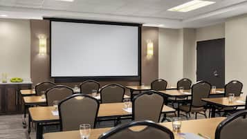 Sala de conferências com várias mesas e cadeiras, uma tela de projetor e iluminação instalada na parede.