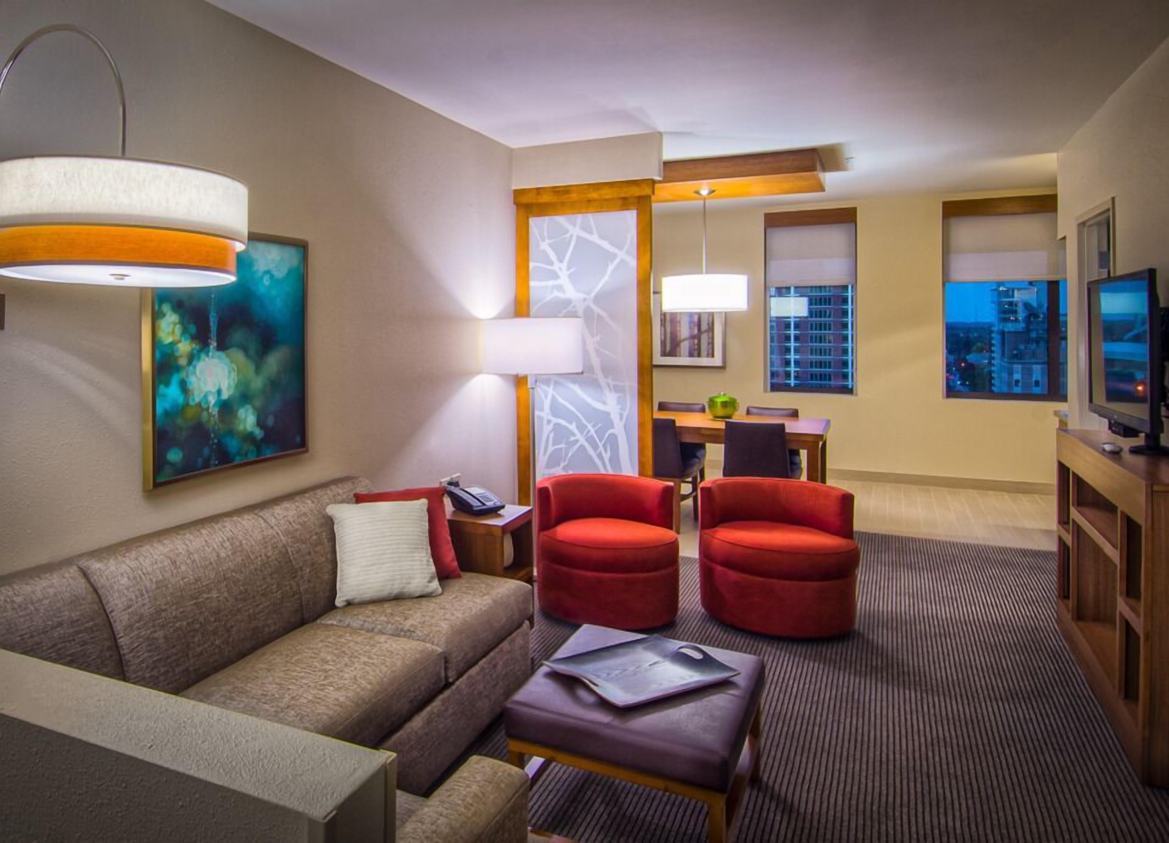 Quarto de hotel com sofá, duas cadeiras vermelhas, uma mesa de jantar com cadeiras, televisão e obras de arte nas paredes.