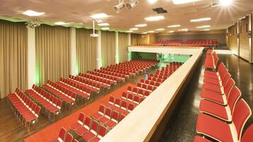 Uma ampla sala de conferências com fileiras de cadeiras vermelhas, uma mesa comprida e um palco com equipamentos de iluminação.