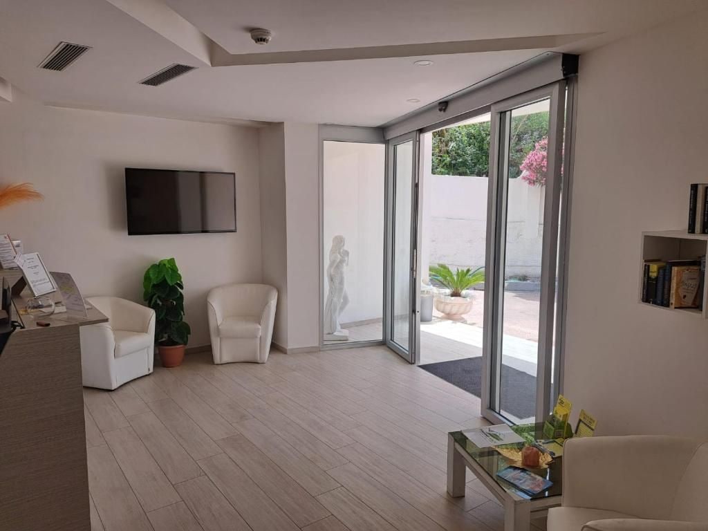 Sala de estar moderna com televisão de tela plana, duas poltronas brancas, uma mesa de centro e uma grande porta de vidro deslizante que dá para uma área externa com plantas.