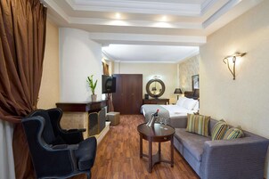 Quarto de hotel com sofá, poltrona, televisão e uma pequena mesa com uma garrafa e copos.