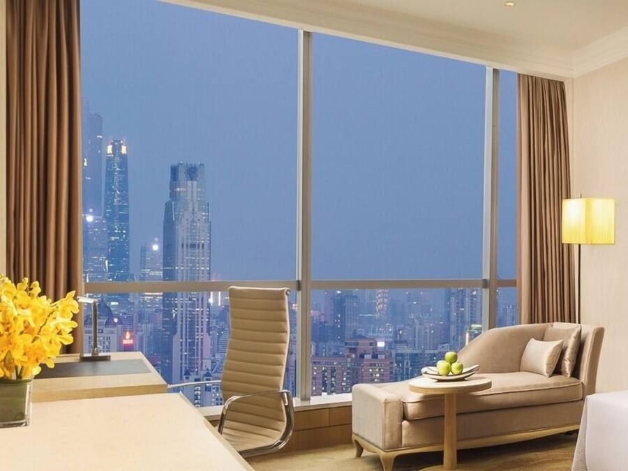 Quarto de hotel com uma janela grande que oferece vista para a cidade, um sofá, uma mesinha com frutas e um vaso de flores.