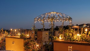 Uma banheira de hidromassagem com estrutura de vidro e luzes de festa, rodeada por plantas e vista para a cidade ao entardecer.