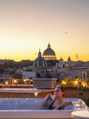 Terraço no telhado com vista para um edifício em forma de cúpula, céu claro e uma pessoa relaxando em uma banheira de hidromassagem.