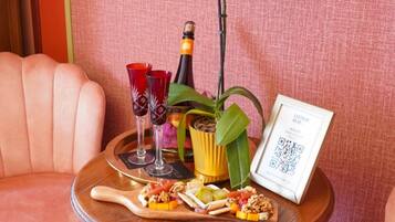 Uma mesa com uma garrafa, copos, uma planta e um código QR em um quarto com cadeiras cor-de-rosa e uma televisão.