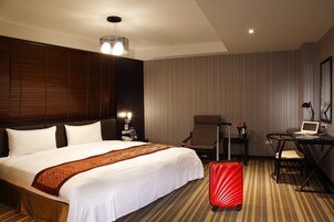 Quarto de hotel com uma cama grande, uma escrivaninha com um laptop, uma cadeira, uma mala vermelha e uma mesinha com um vaso de flores.