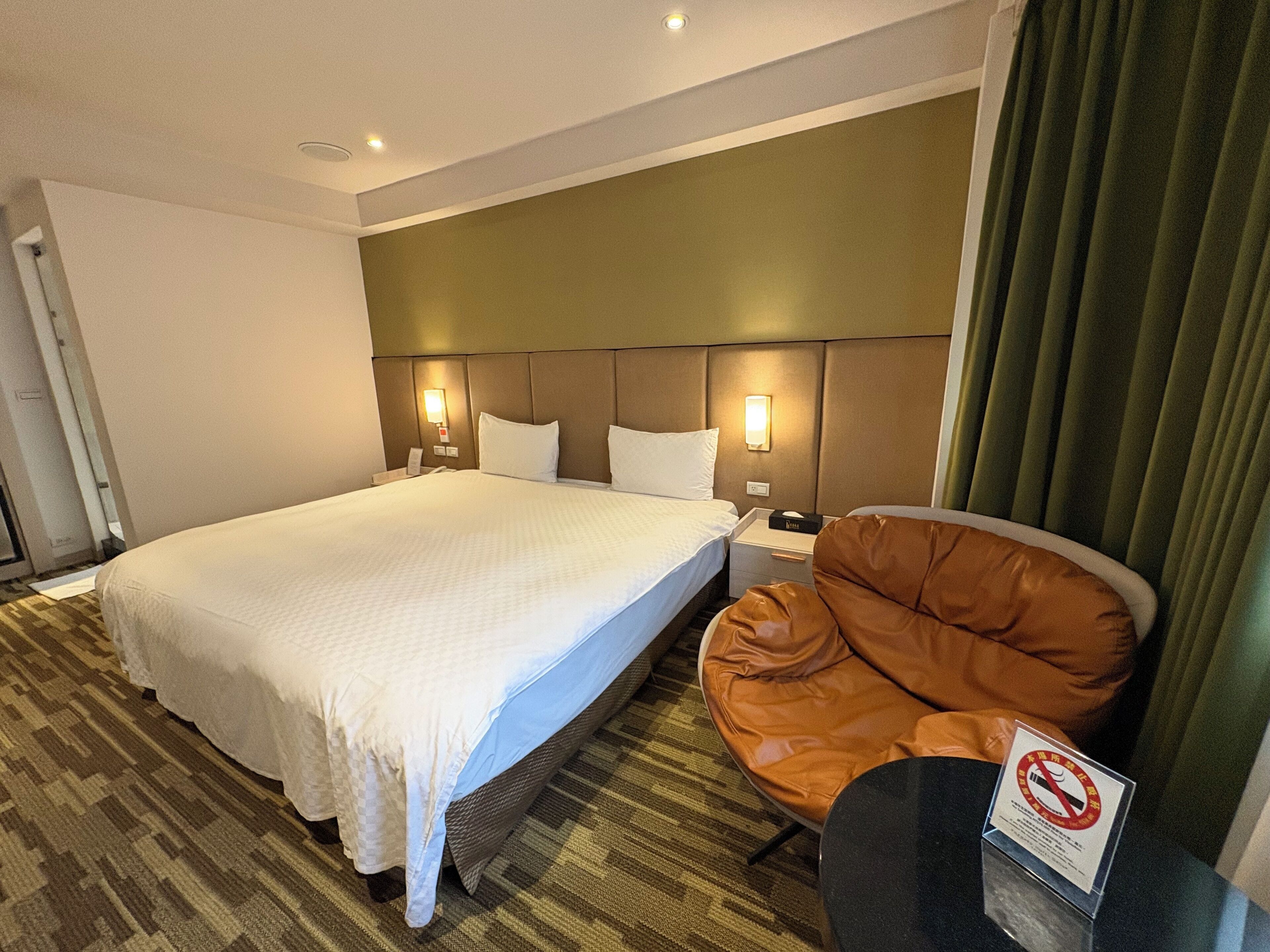 Quarto de hotel com uma cama grande, uma poltrona de couro marrom, uma mesinha de cabeceira e um aviso de proibição de fumar.