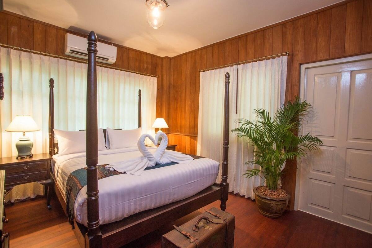 Uma cama de dossel com arranjo de toalhas em formato de coração, um vaso de planta e uma mesa de cabeceira de madeira com abajur.