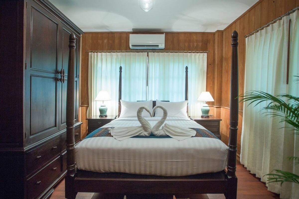 Uma cama bem arrumada com arranjo de toalhas em formato de coração, painéis de madeira e duas luminárias de cabeceira.
