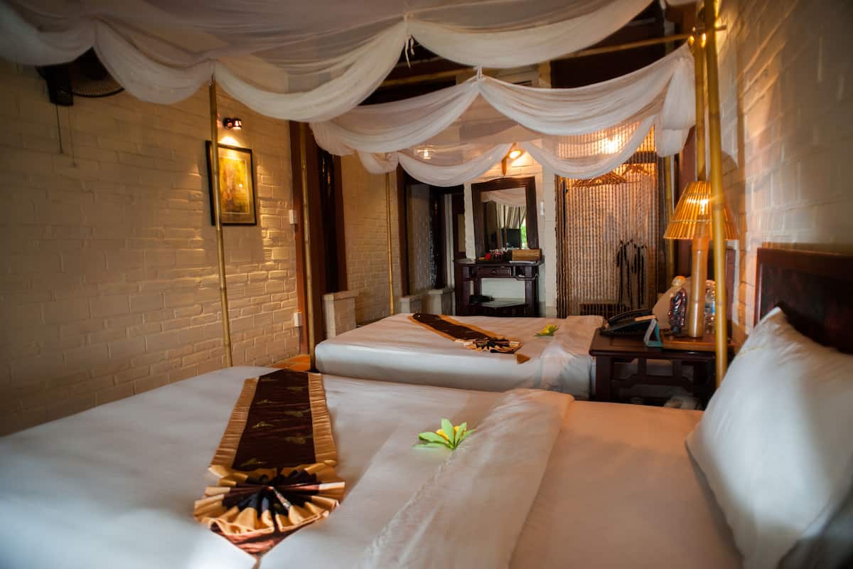Quarto de hotel com duas camas, cortinas brancas, um espelho, mesa de cabeceira com abajur e um quadro na parede.