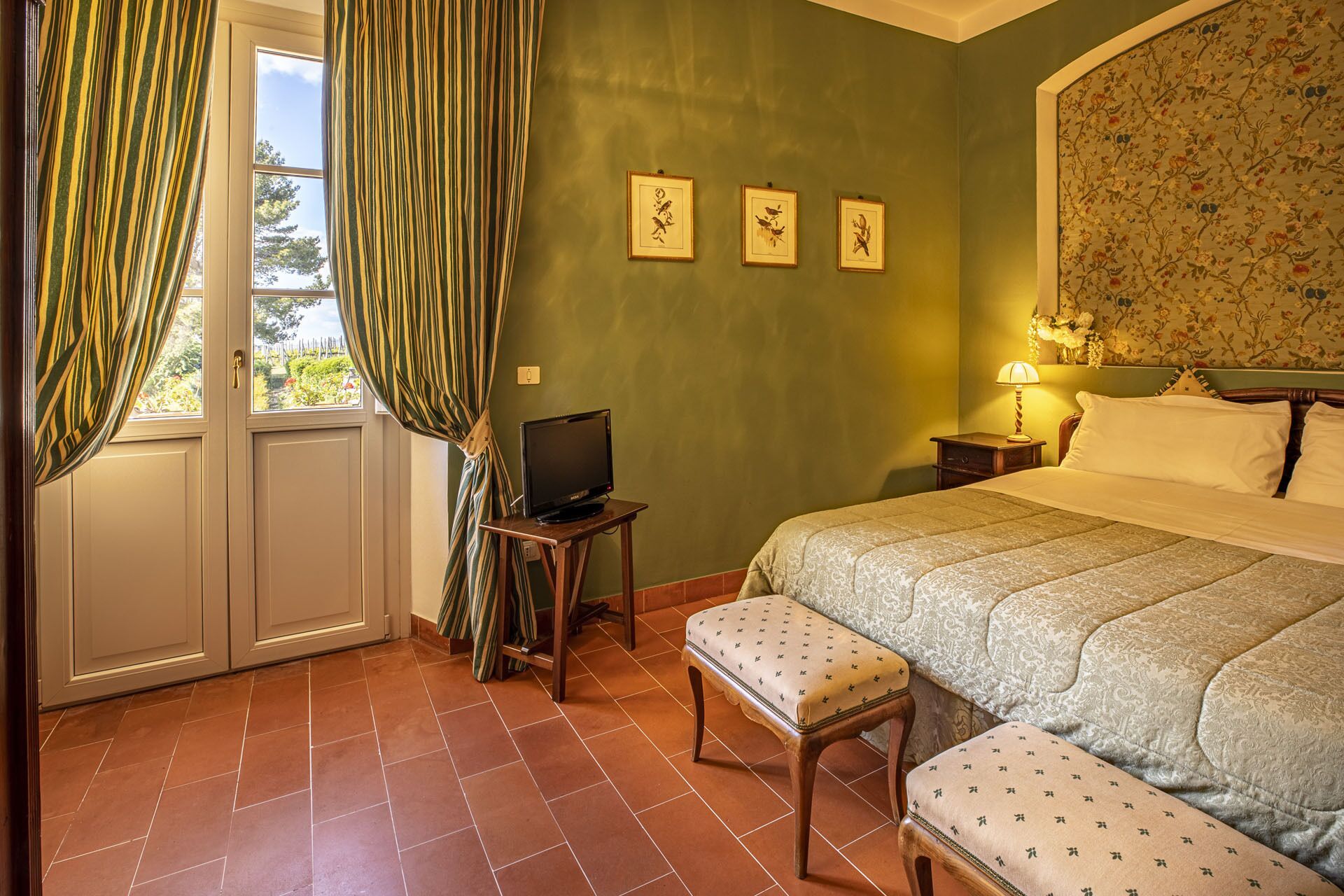 Quarto de hotel com uma cama, televisão, um banco, mesa de cabeceira com abajur e três quadros na parede.