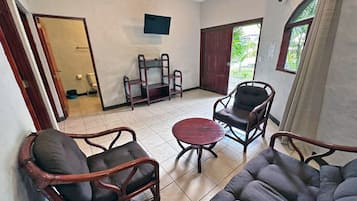 Quarto com sofá, duas cadeiras, uma mesa pequena, televisão e porta para o banheiro.