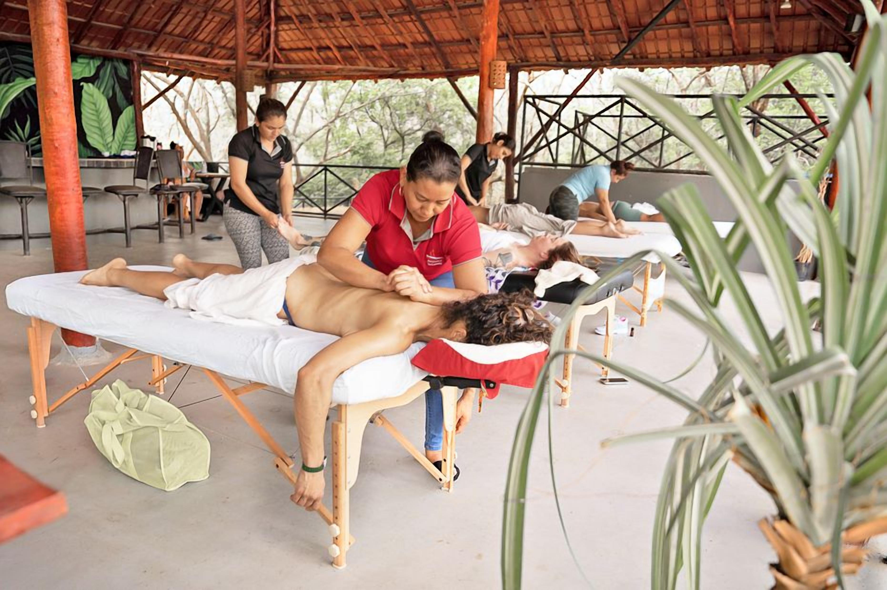 Sessão de massagem em área externa coberta, com bancos de madeira e telhado de palha.