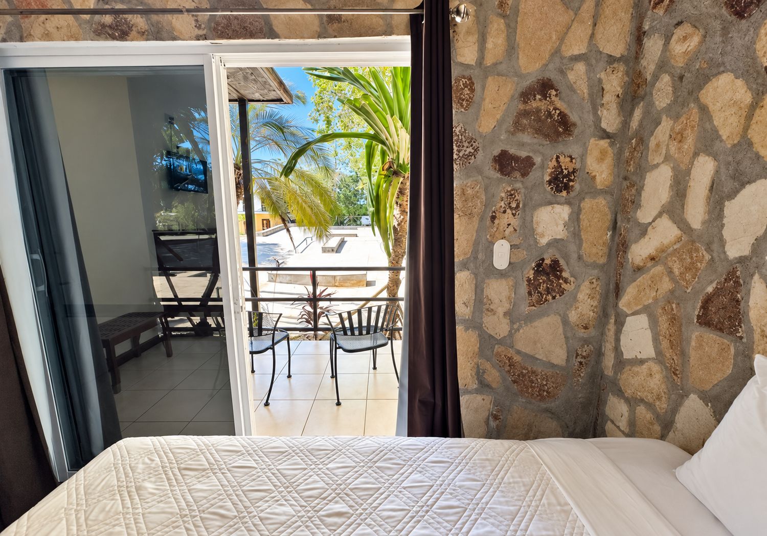 Quarto com uma cama, vista para um pátio externo com cadeiras e uma mesa, e uma parede de pedra.