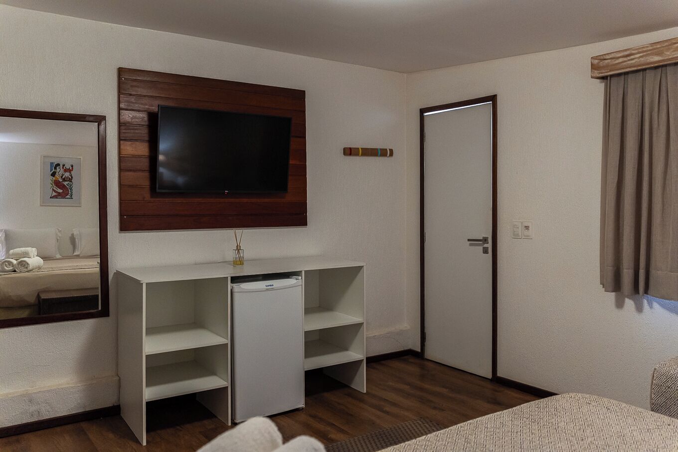 Quarto de hotel compacto com televisão de tela plana fixada em um painel de madeira, guarda-roupa branco com espelho, porta com puxador e janela com cortinas.