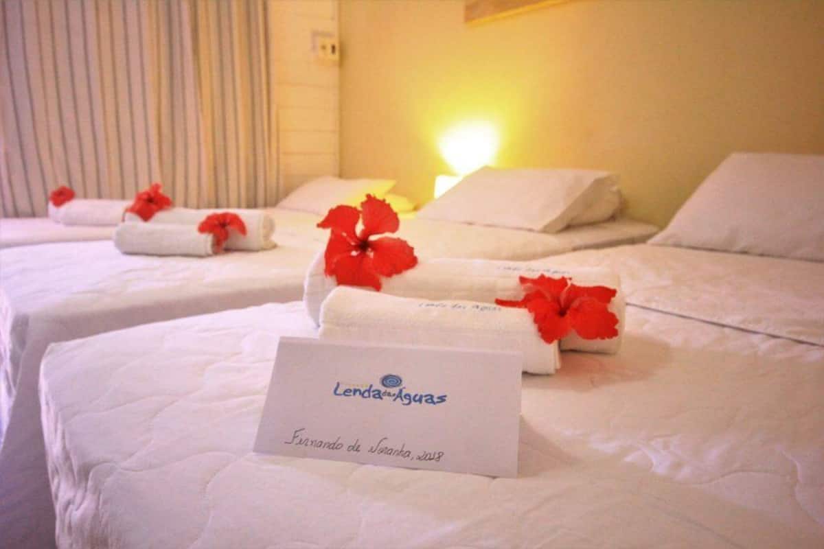 Quarto de hotel com camas arrumadas, cada uma com travesseiro branco e uma flor vermelha. Uma placa na cama diz 'Lenda de Agua'.