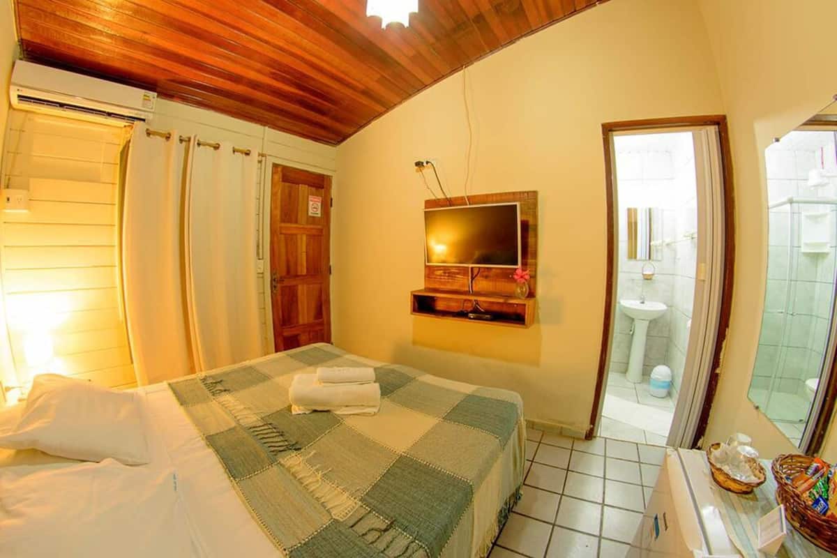 Quarto com cama, teto de madeira, televisão montada e banheiro visível através de uma porta aberta.