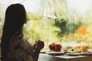 Uma pessoa sentada à mesa com uma refeição, um copo de suco e uma janela com vista para o verde.