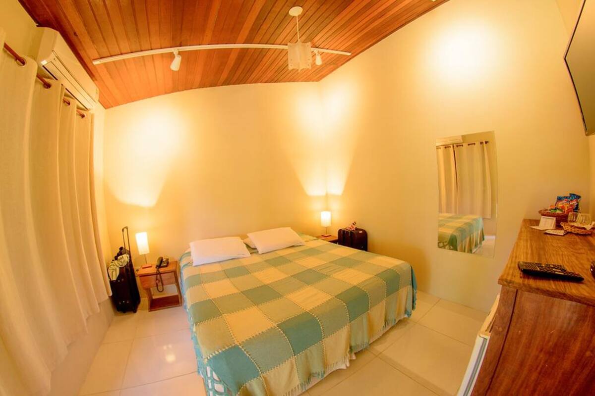 Quarto de hotel com colcha xadrez, teto de madeira, mesa de cabeceira com abajur, espelho e escrivaninha com teclado e mouse.