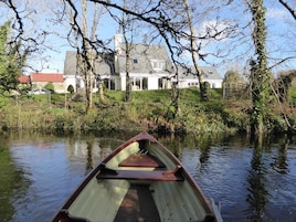 Um canal estreito com um barco a remo em primeiro plano, uma casa com telhado vermelho e paredes de pedra ao fundo, e uma área gramada com árvores e arbustos.