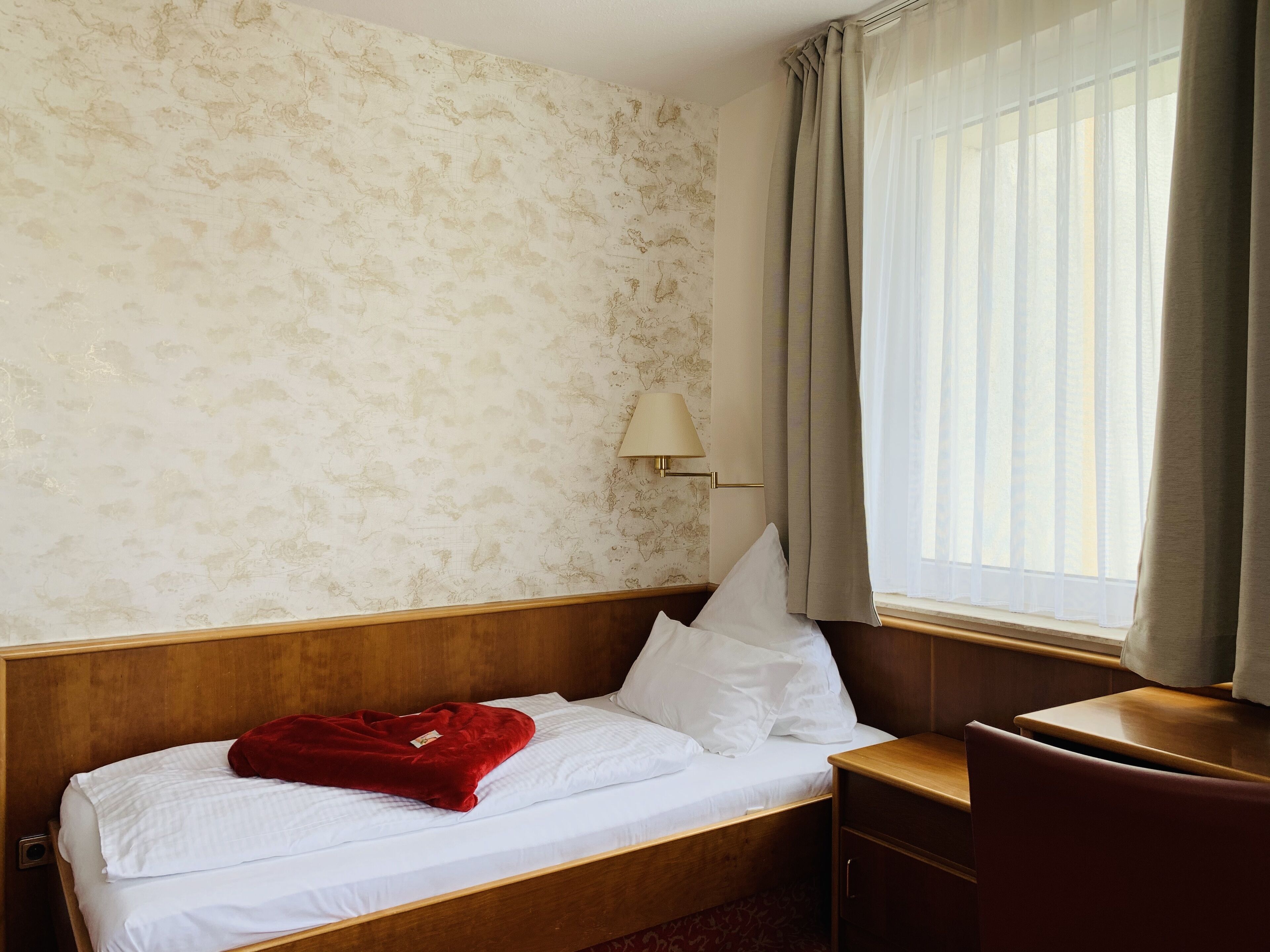 Quarto de hotel com cabeceira de madeira, cama com lençóis brancos, manta vermelha, mesa de cabeceira, abajur e janela com cortinas finas.