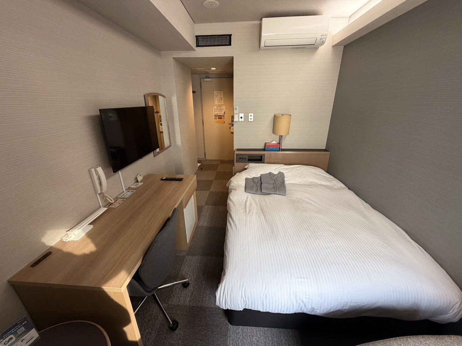 Quarto de hotel com cama, secretária com cadeira, televisão, ar condicionado e luminária.