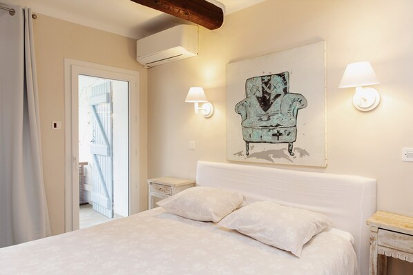 Quarto com cama, duas mesas de cabeceira, uma luminária na parede e uma porta que dá para outro cômodo.