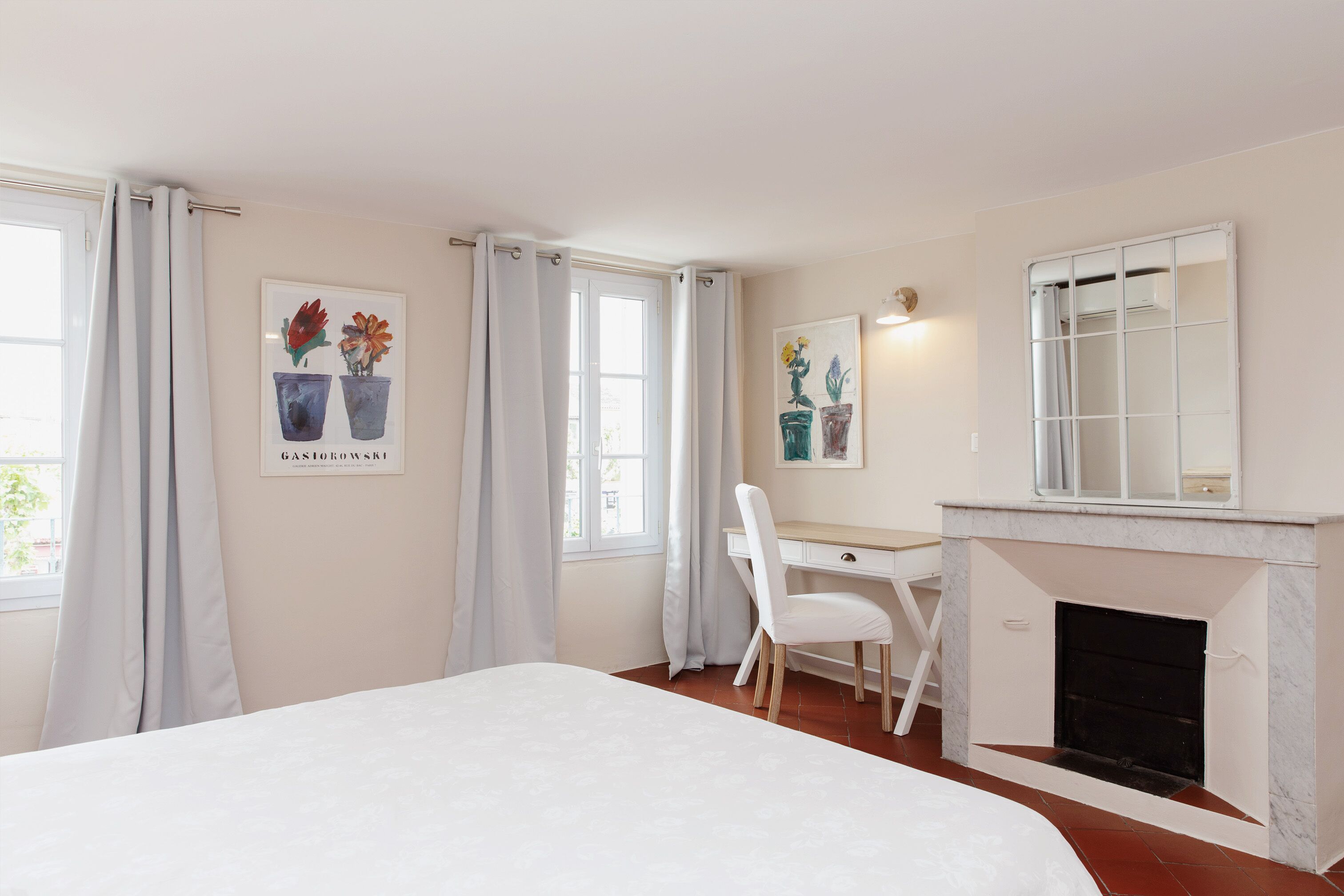Quarto com cama, escrivaninha, cadeira, lareira e duas janelas com cortinas.