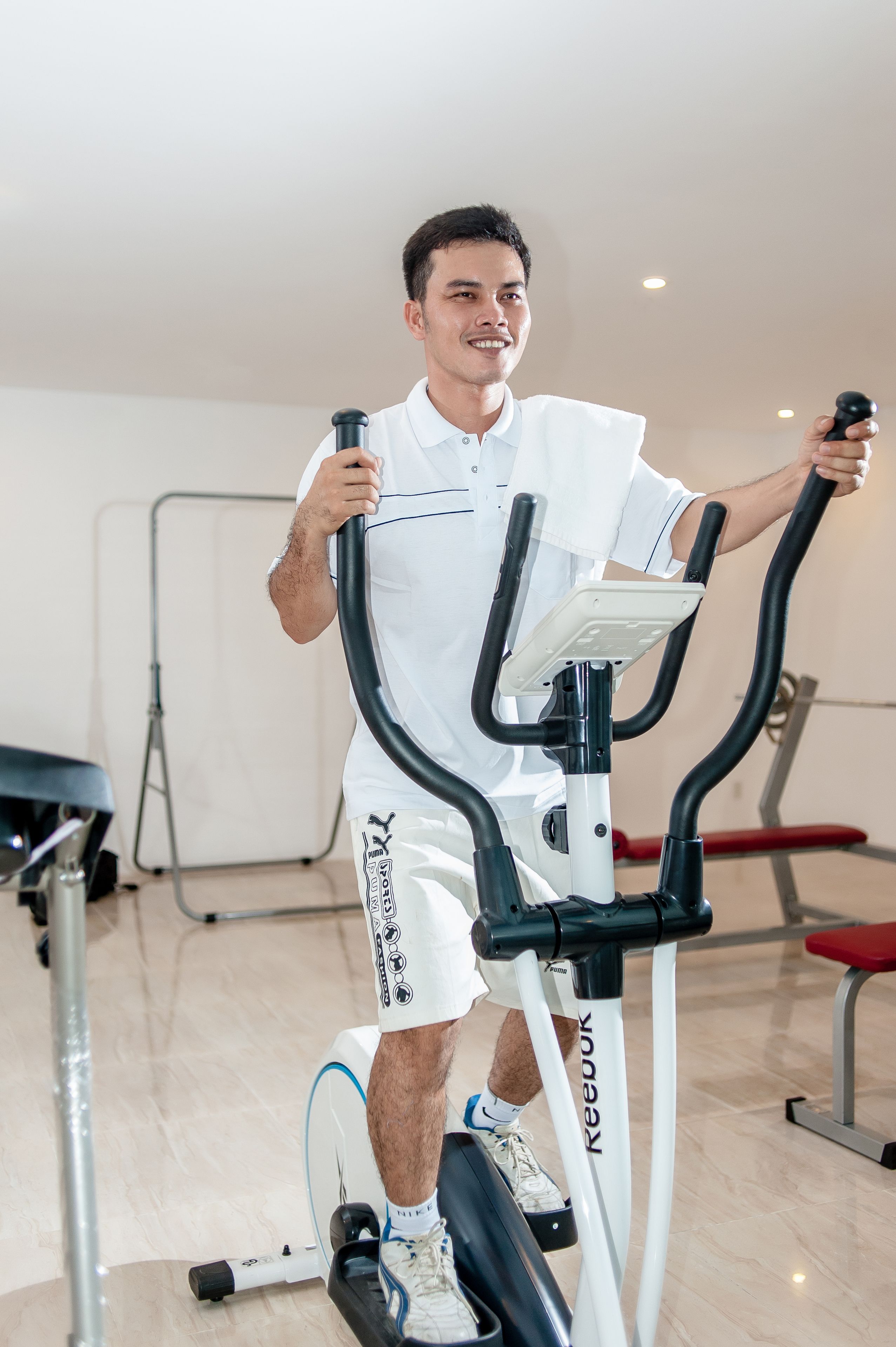 Uma pessoa usando um elíptico numa academia, com uma esteira e outros equipamentos de exercício ao fundo.
