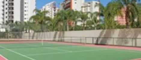 Uma quadra de tênis ao ar livre, com superfície de jogo verde e área circundante vermelha, cercada por edifícios e palmeiras.