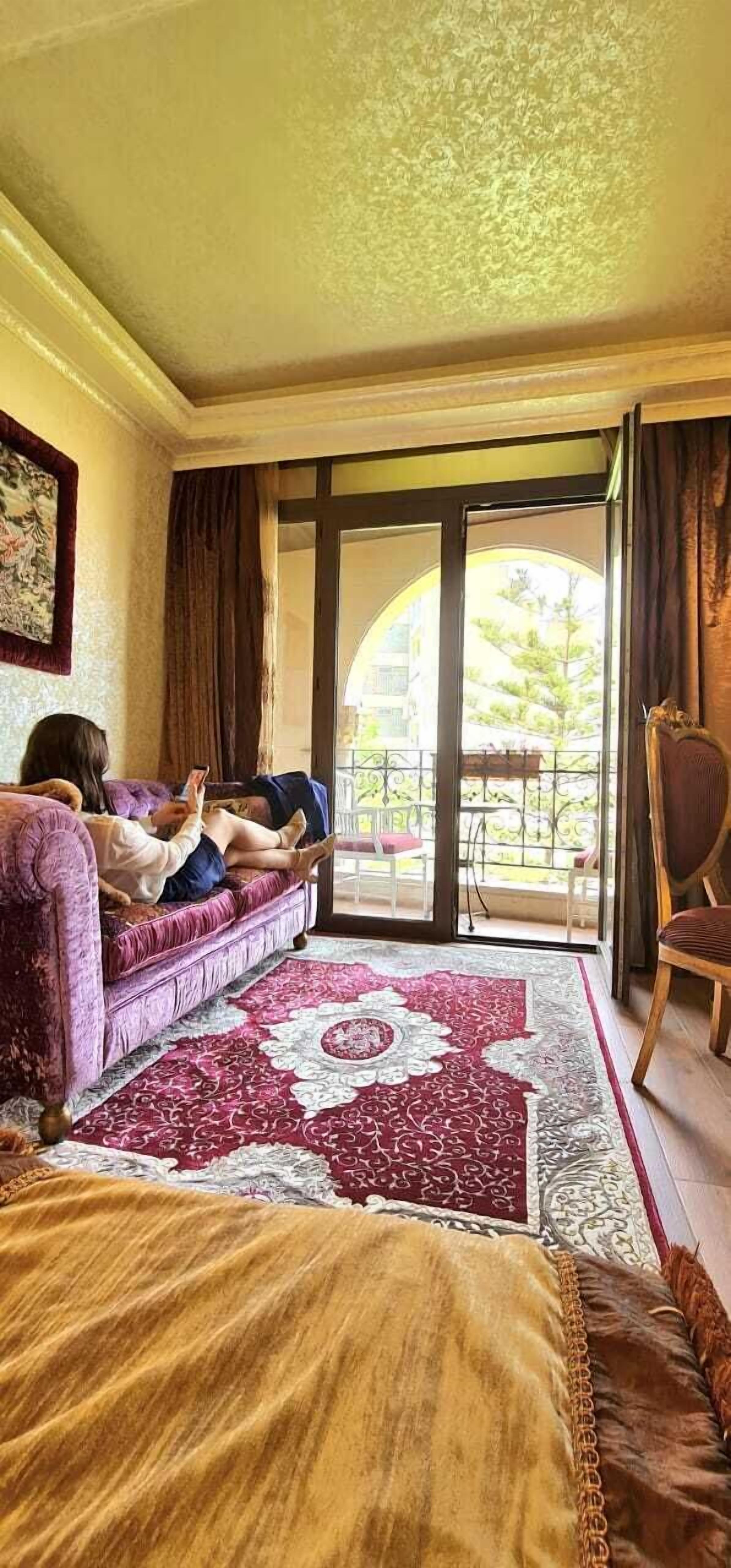 Quarto com sofá roxo, cadeira de madeira e tapete estampado.