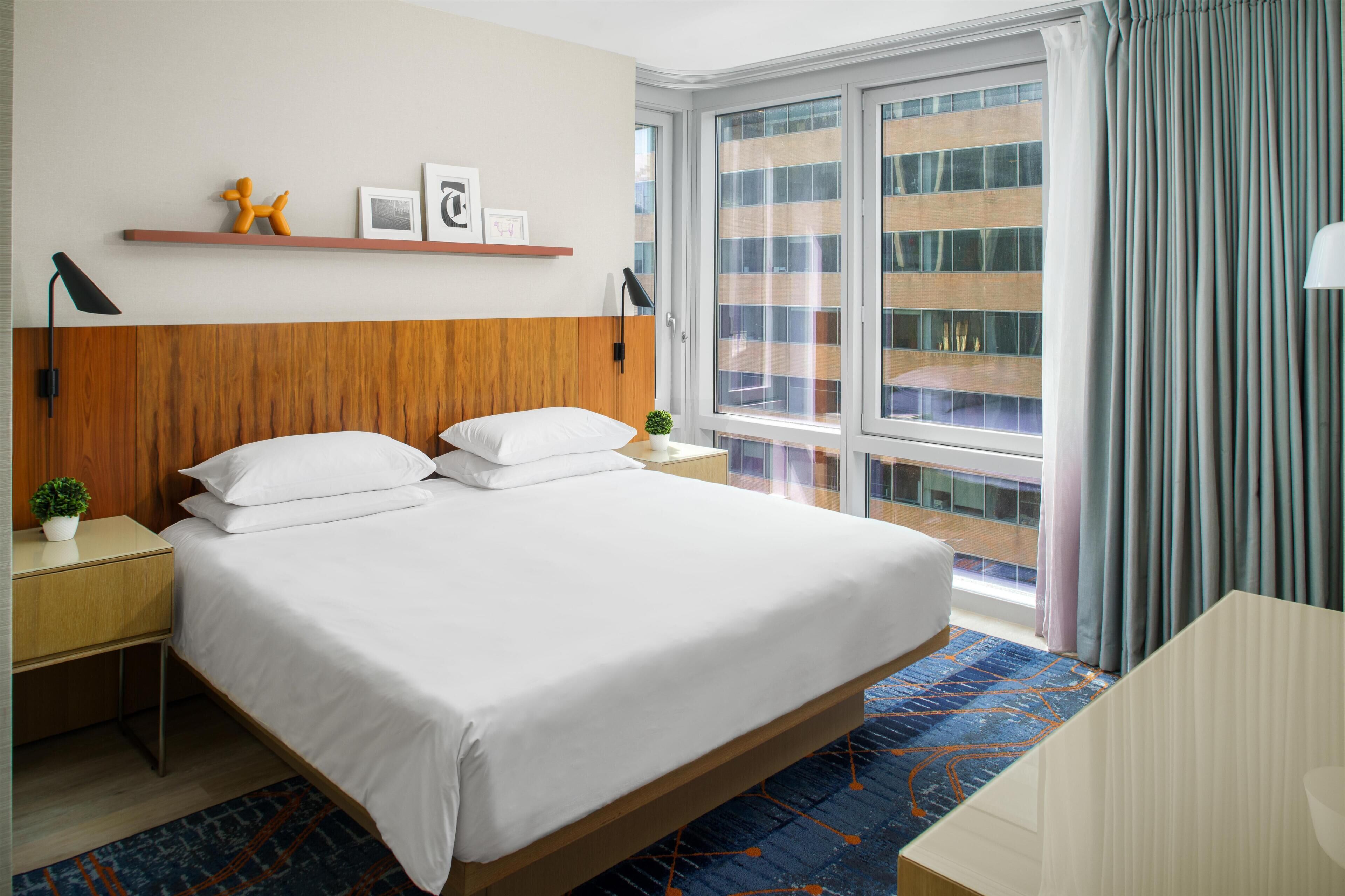 Quarto de hotel com cama grande, mesa de cabeceira, escrivaninha, janela com cortinas e vista para o edifício.