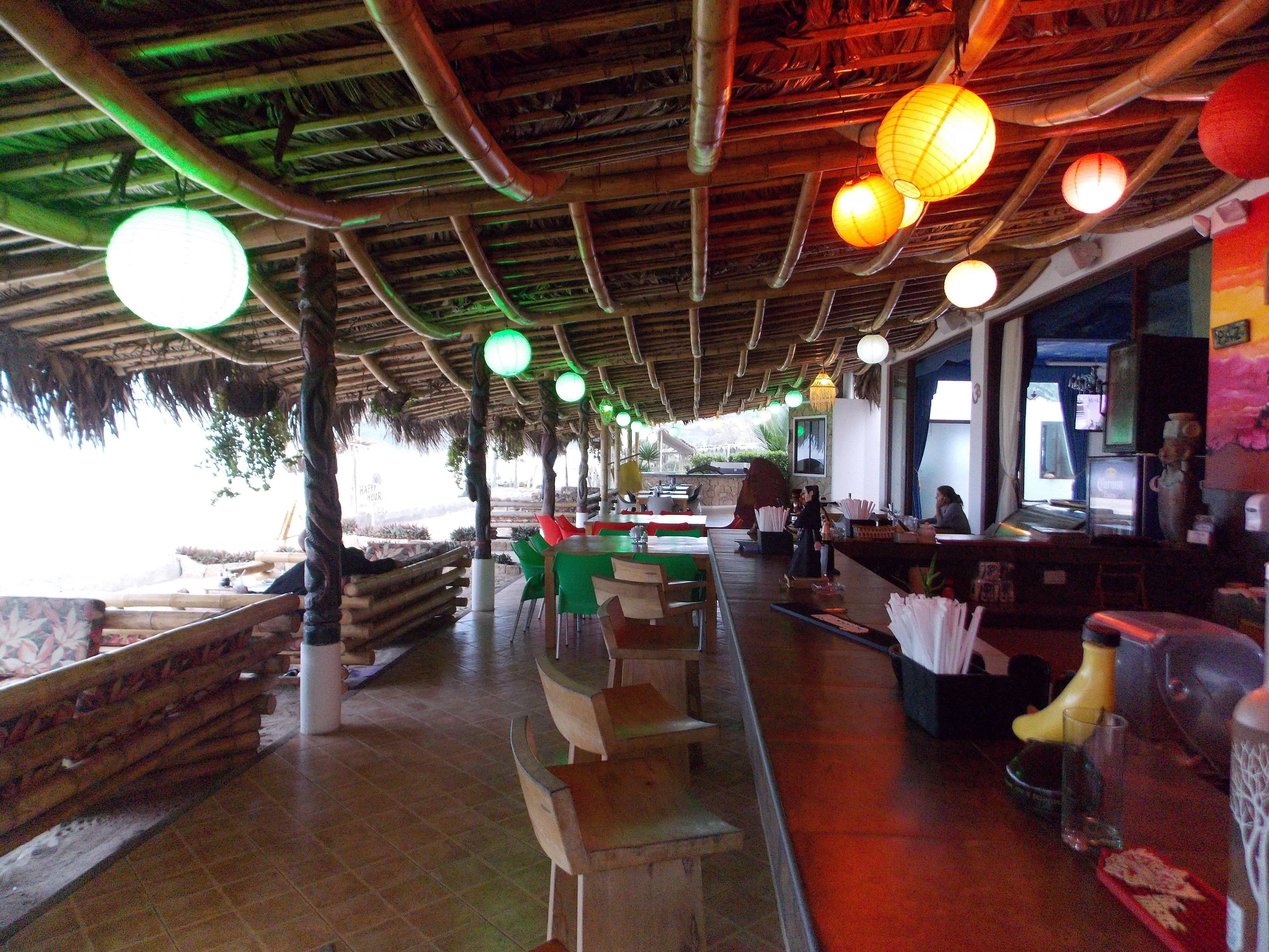 Restaurante com telhado de palha, luminárias pendentes verdes e laranja, e vista para a praia.