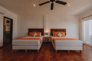 Quarto de hotel com duas camas, um ventilador de teto, uma pequena mesa e uma janela grande.