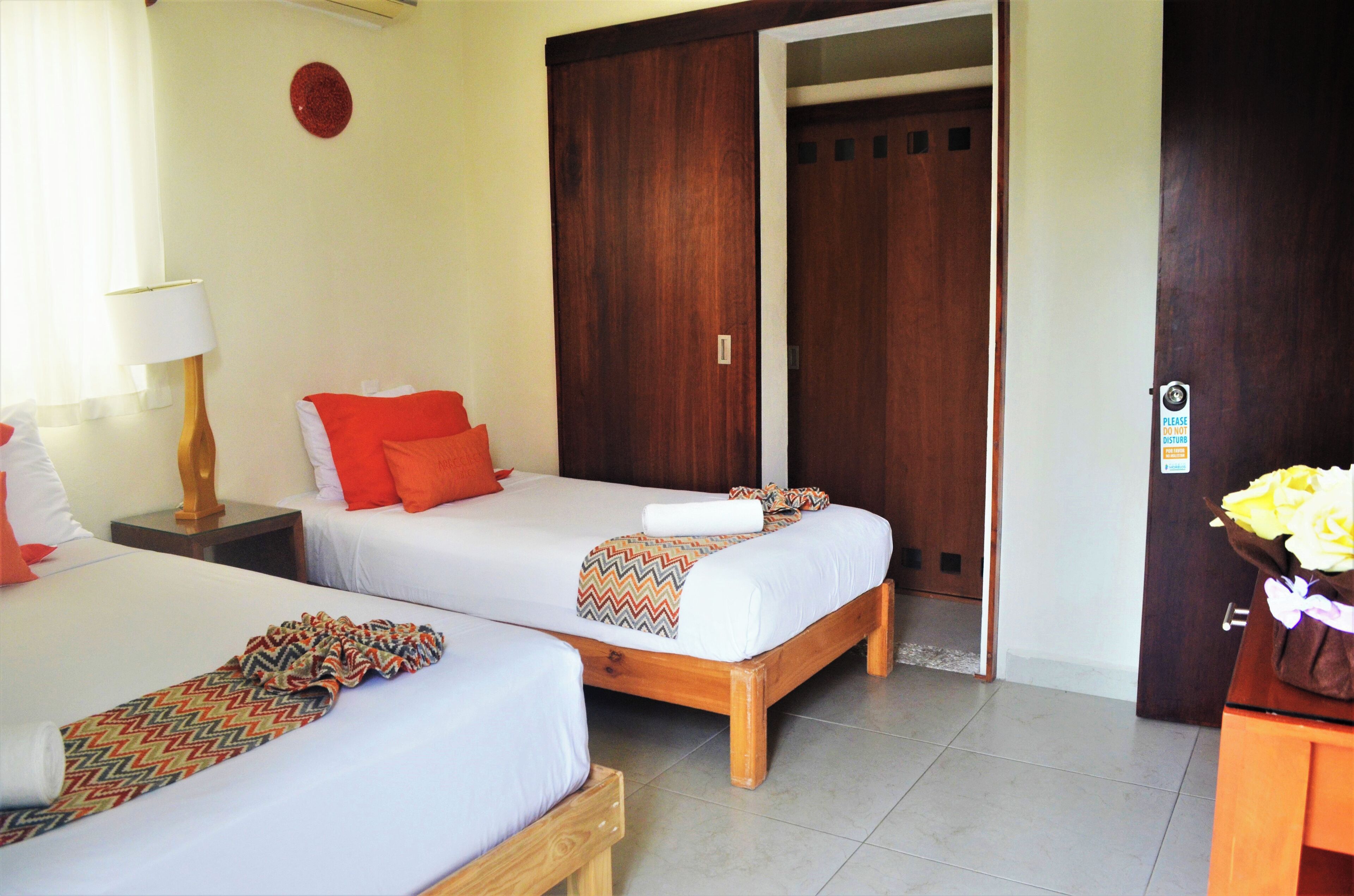 Quarto de hotel com duas camas de solteiro, um guarda-roupa de madeira, uma mesa de cabeceira com abajur e uma porta com a placa 'Não Perturbe'.