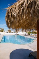 Piscina com cobertura de palha, cadeiras de sol e um céu azul límpido.