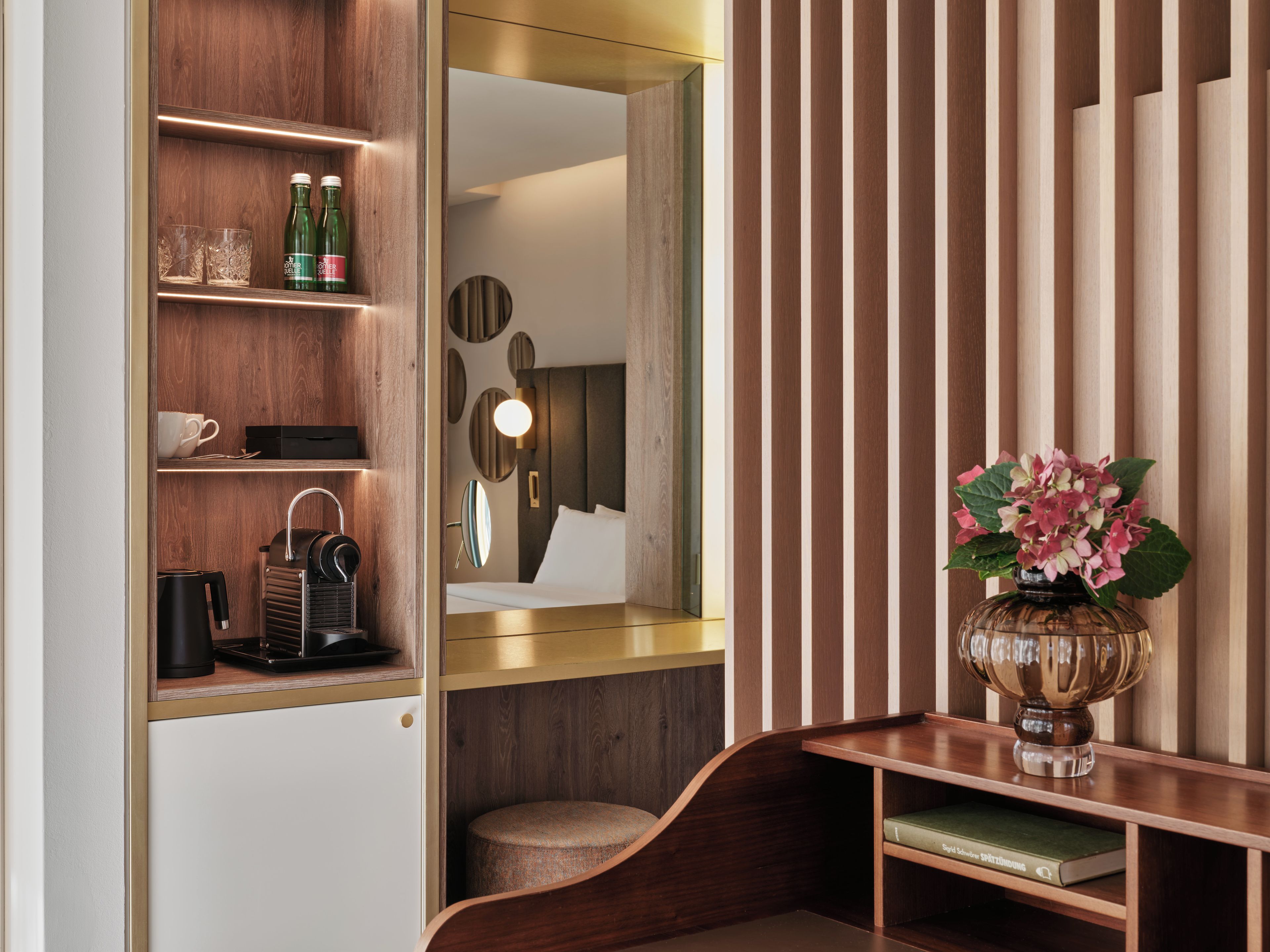 Quarto de hotel moderno com toucador de madeira, espelho, mesa de cabeceira com um vaso de flores e área de estar com um banco.