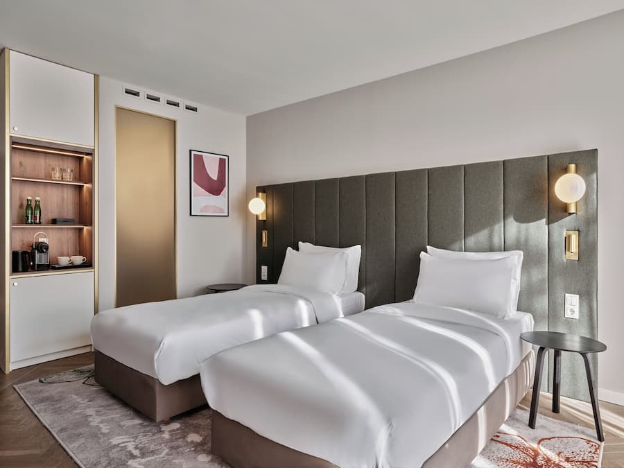 Quarto de hotel com duas camas, um minibar, uma luminária fixada na parede e uma obra de arte emoldurada.