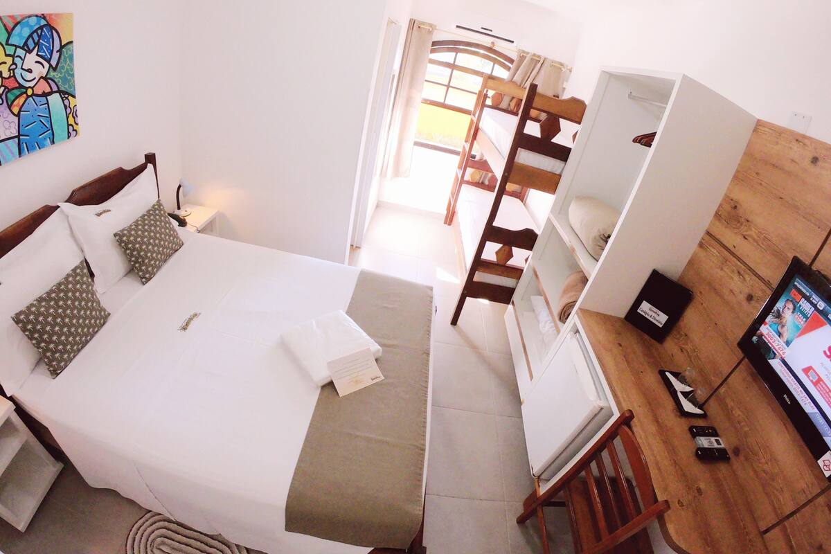 Quarto de hotel com cama, escrivaninha, cadeira, televisão e piso de madeira.