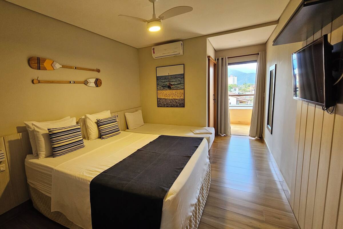 Quarto de hotel com cama, ventoinha no teto, aparelho de ar condicionado, um quadro e vista para o exterior.