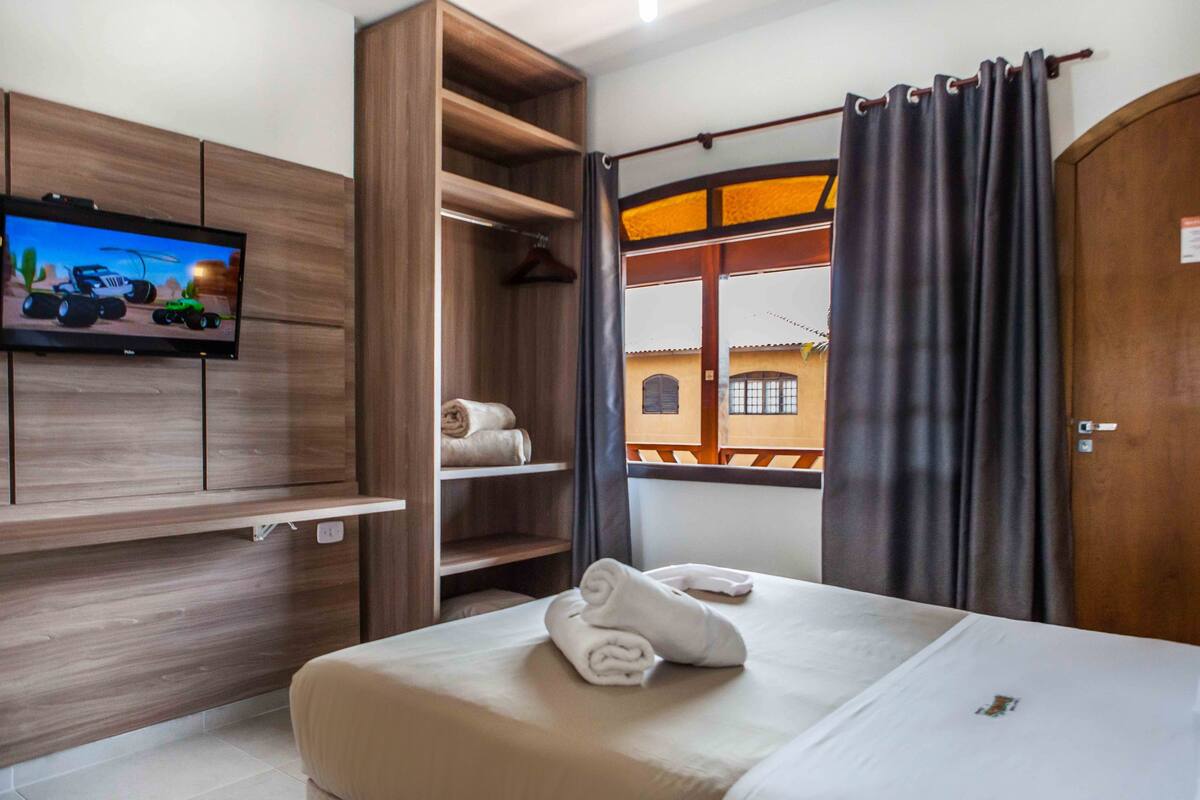 Quarto de hotel com uma cama, televisão presa a um móvel de madeira, guarda-roupa e janela com cortinas.