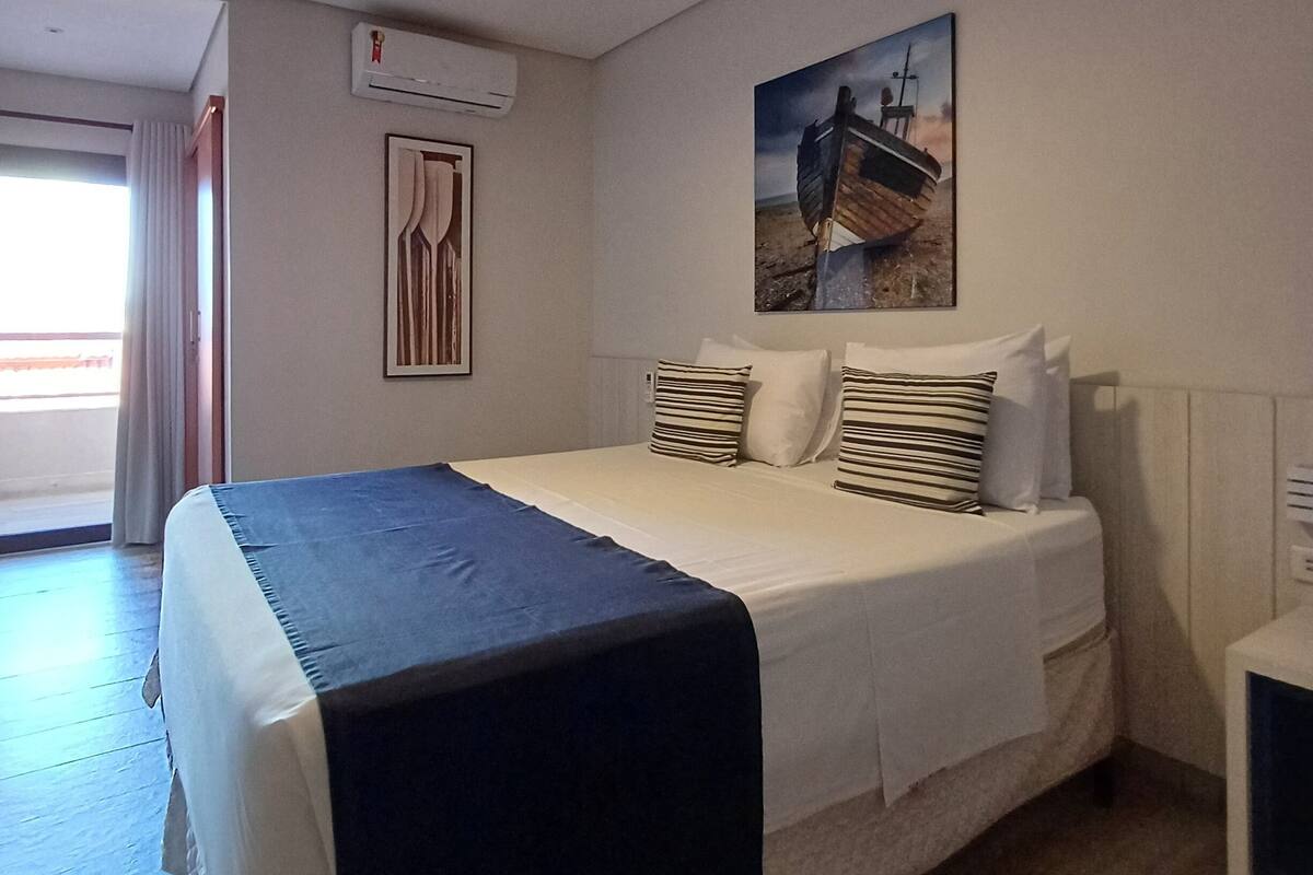 Quarto de hotel com cama, ventilador de teto, ar condicionado, um quadro na parede e janela com cortinas.