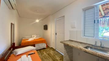 Quarto de hotel compacto com duas camas, uma kitchenette pequena, uma janela com persianas e um aparelho de ar condicionado.