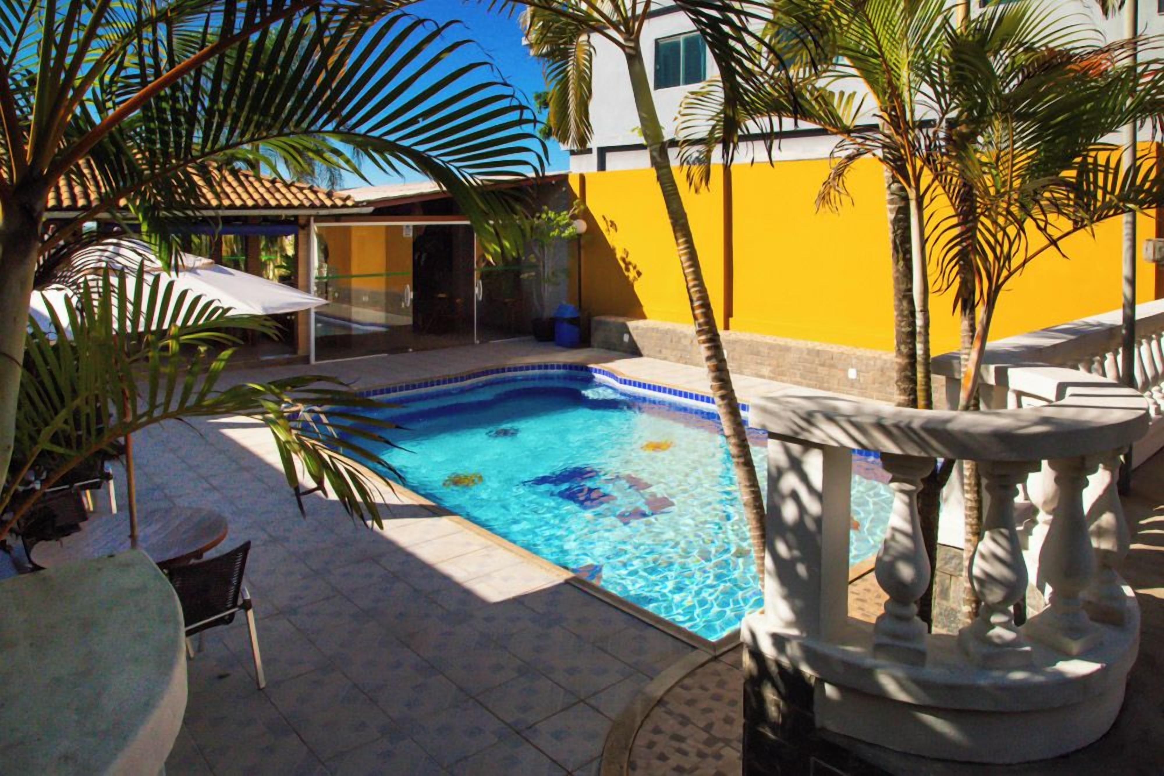 Piscina com deck ladrilhado, rodeada por palmeiras e um prédio com paredes amarelas.